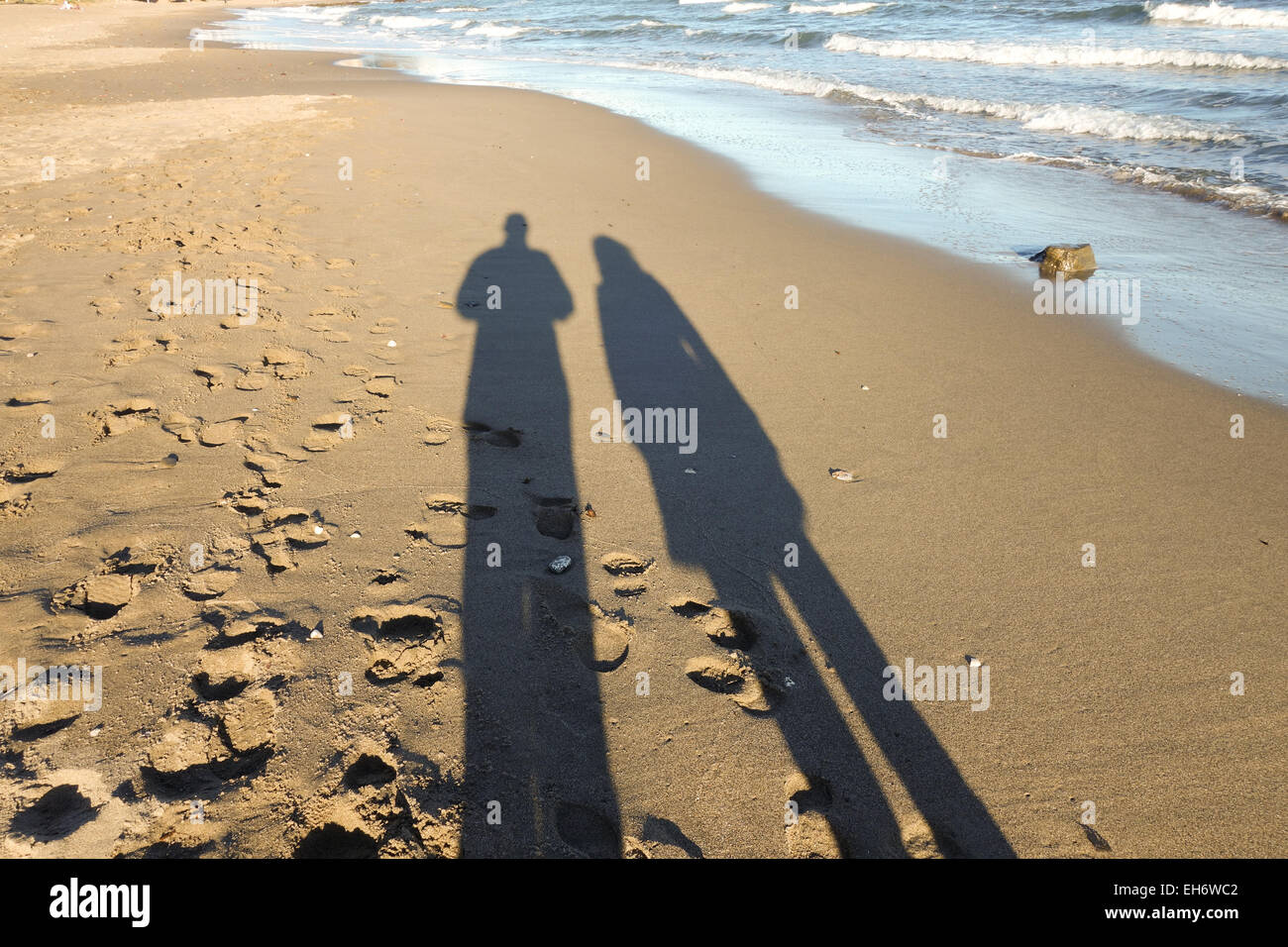 Long Shadow of couple on beach with sea and waves behind at sunset ...