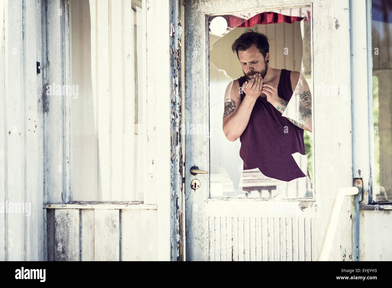 Cool young man smoke cigarette behind broken door and window inside ...