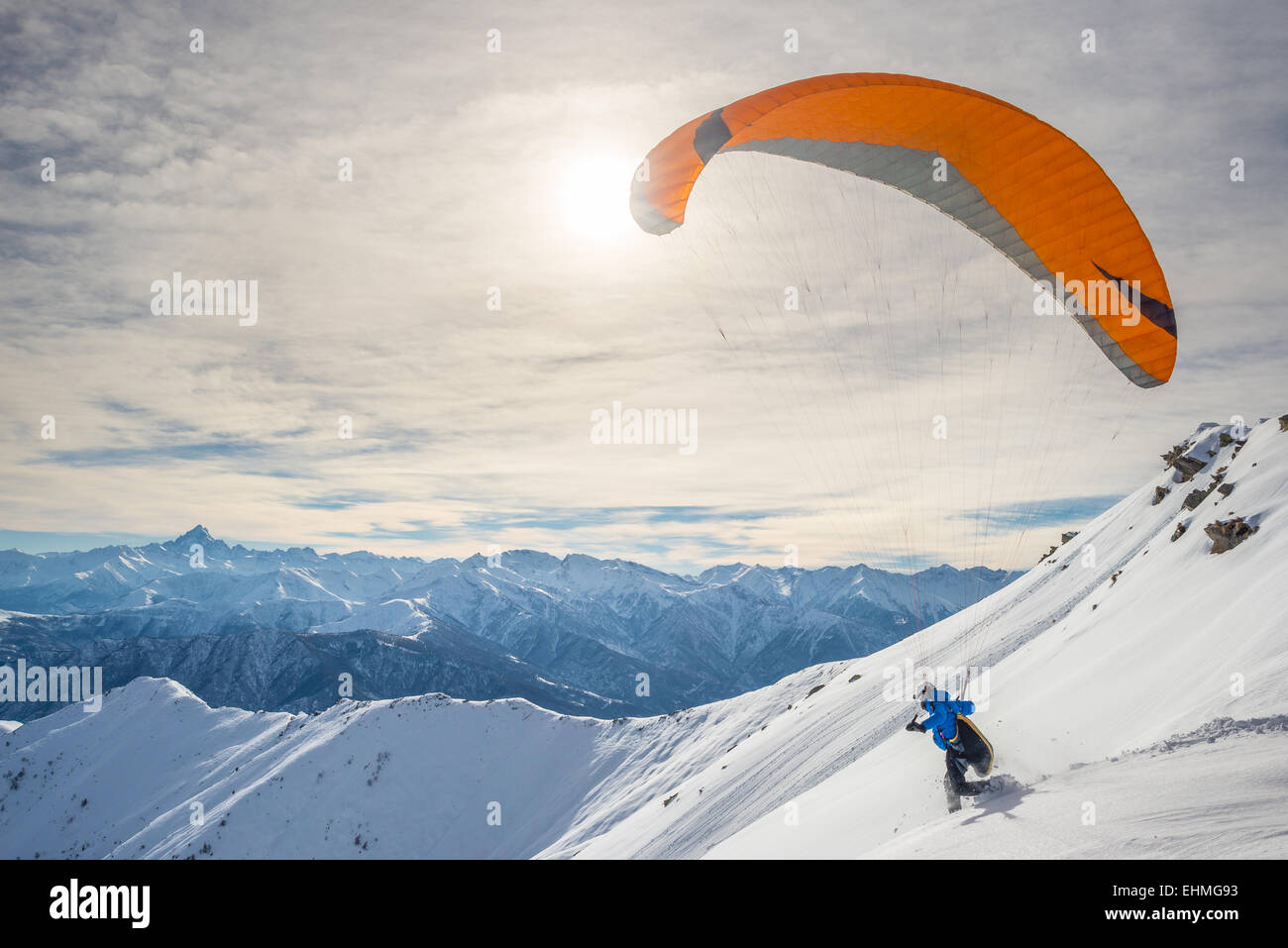 Paraglider running on snowy slope for take off with bright orange kite ...