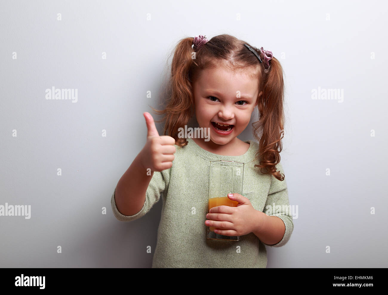 Laughing kid girl drinking juice and showing thumbs up sign on blue ...