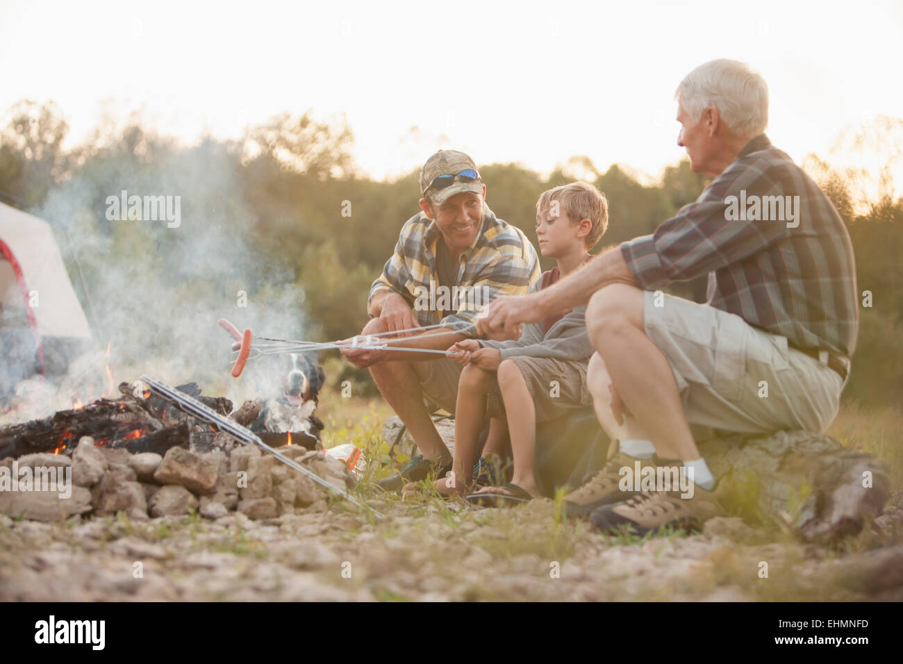 Three generations of Caucasian men roasting hot dogs over campfire ...