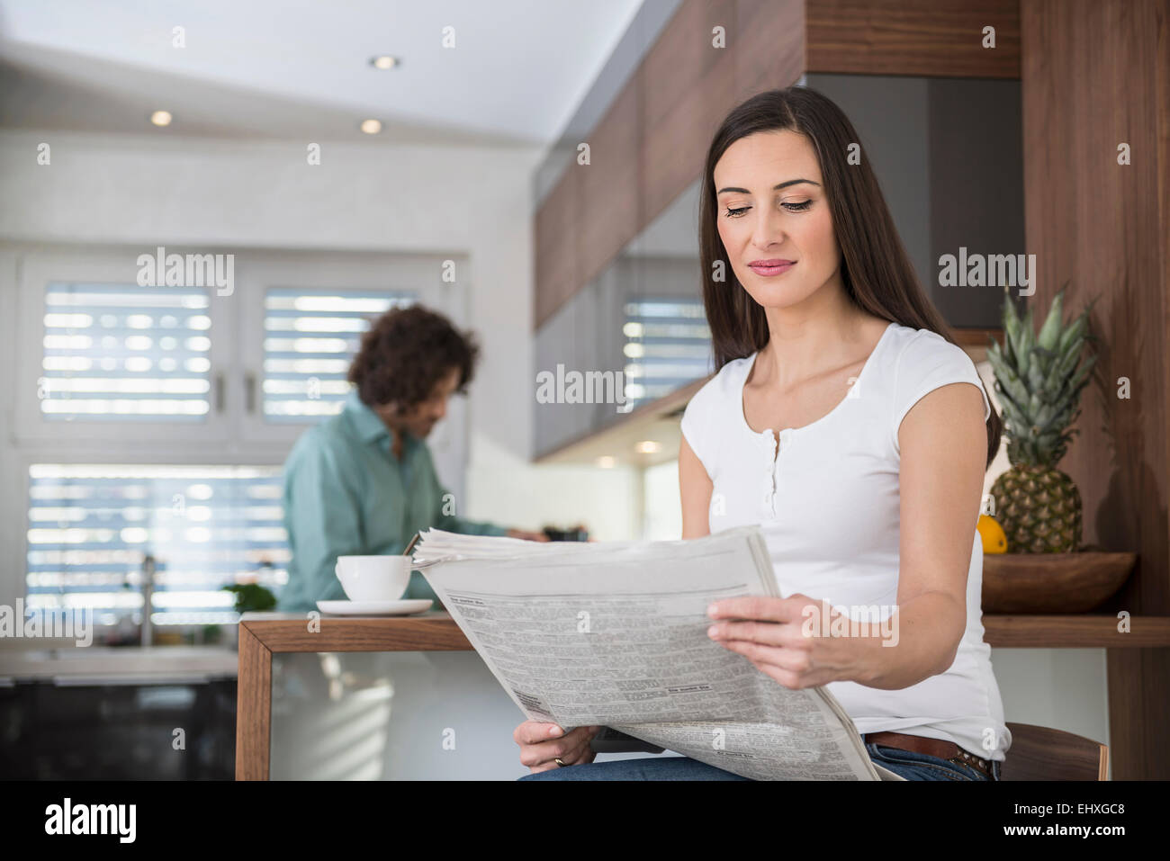 Woman reading a newspaper while her husband cooking in a kitchen ...