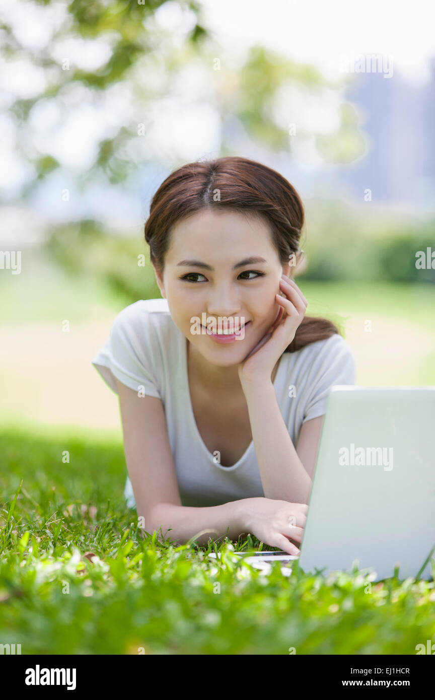 Young woman lying down on front with laptop and looking away with smile ...