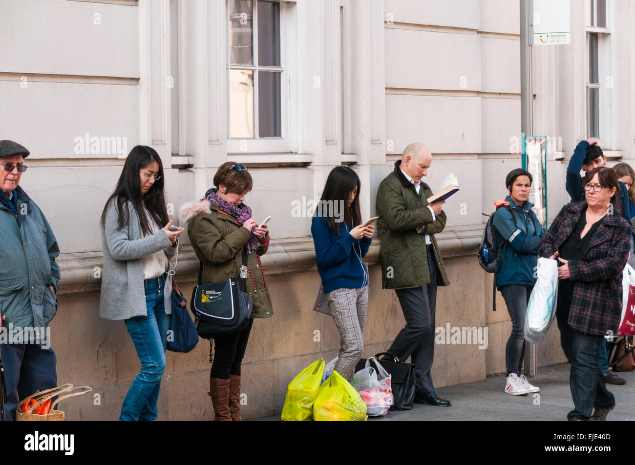 A queue of people standing at a bus stop either reading or checking ...