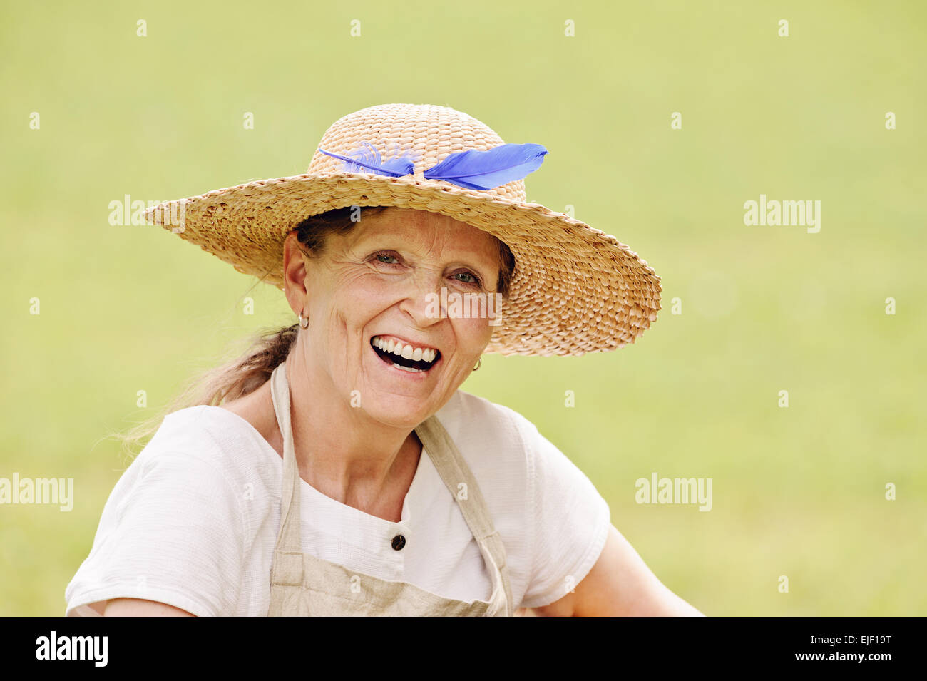 Senior woman wearing a straw hat is looking at teh camera and laughing. The background is a ...