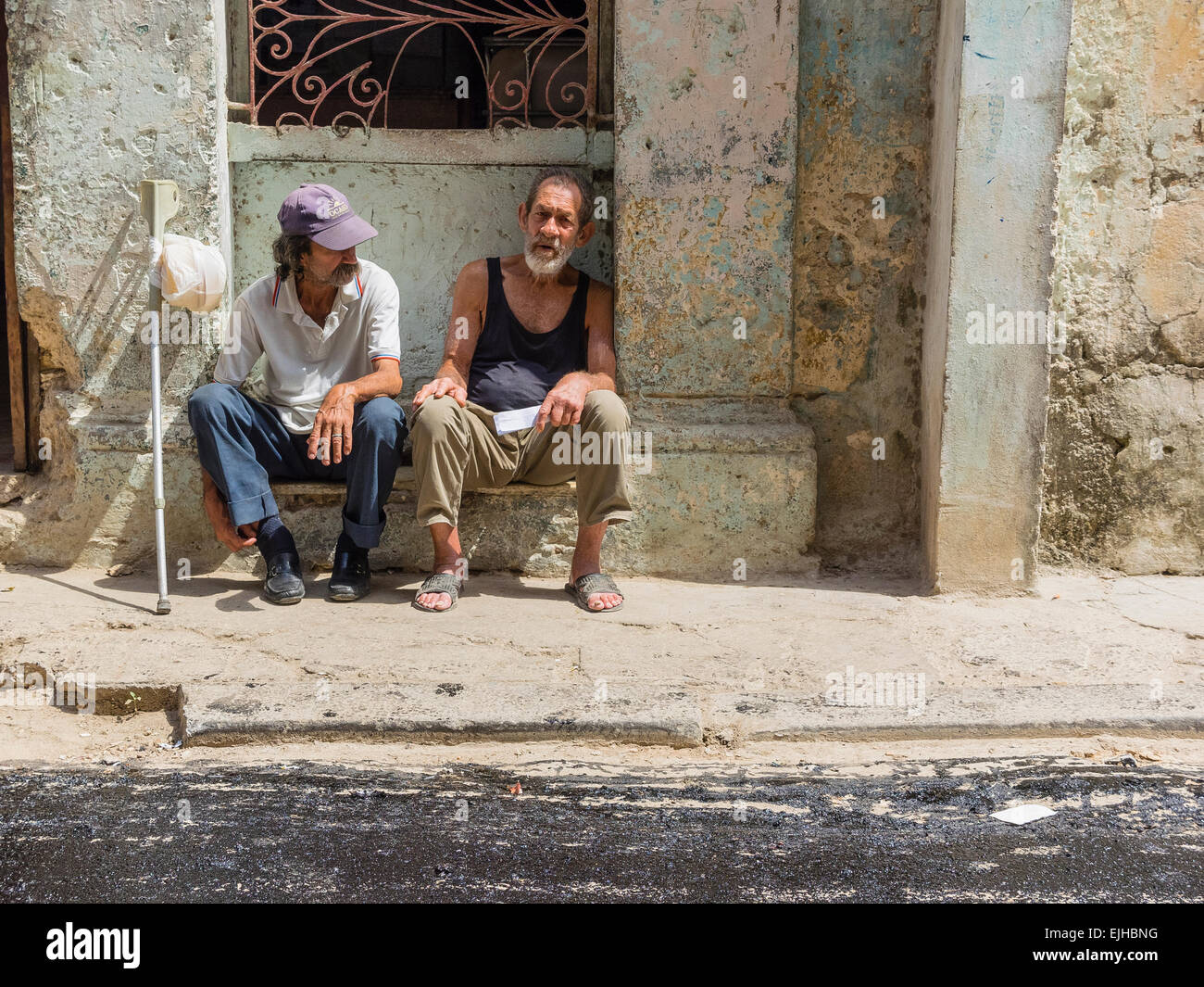 Two Cuban Hispanic male seniors sit on a concrete bench in front of a ...
