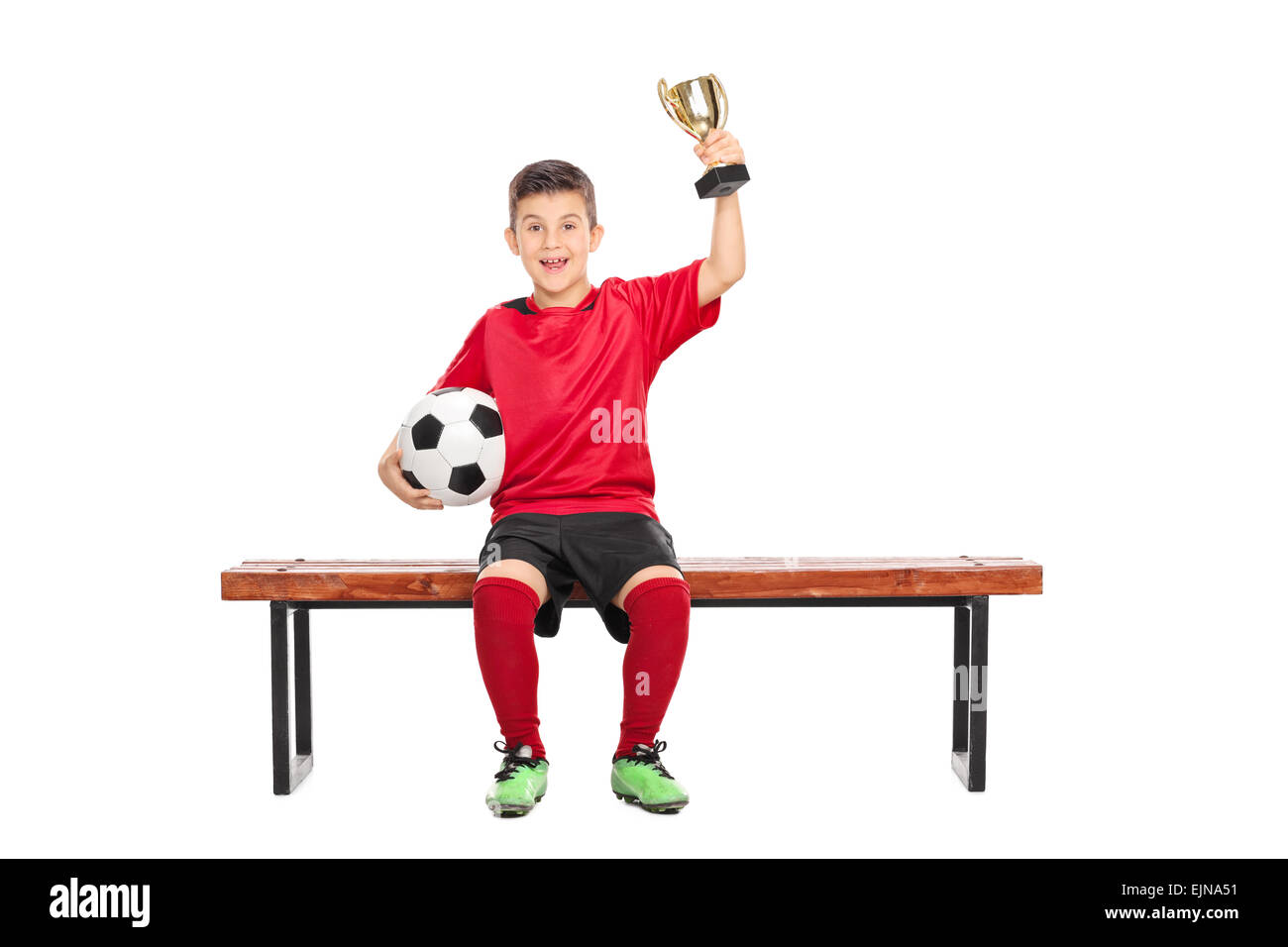 Delighted boy in soccer uniform holding a trophy in one hand and a ball ...