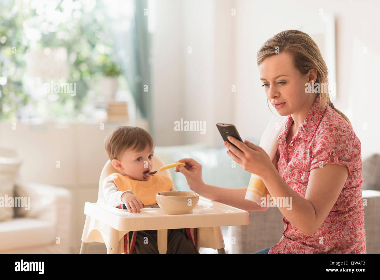Mother feeding little boy (2-3 years) in high chair and texting message ...