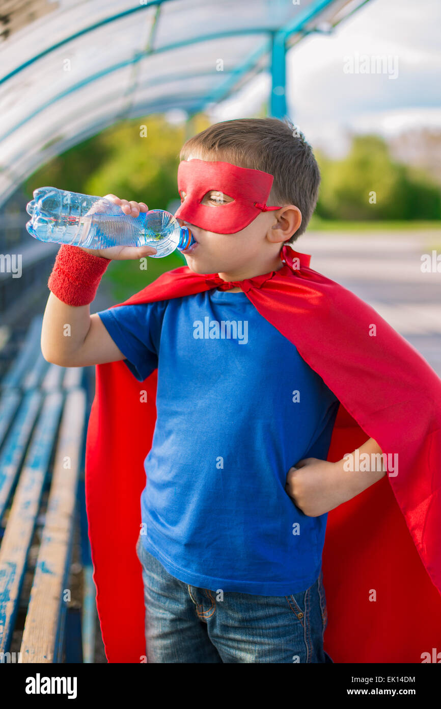 Boy dressed in cape and mask standing and drinking water from a bottle ...