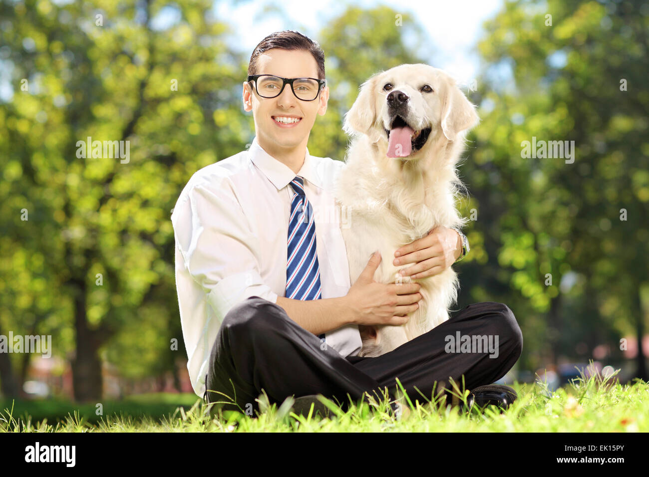 Young smiling man sitting on a green grass and hugging his labrador ...