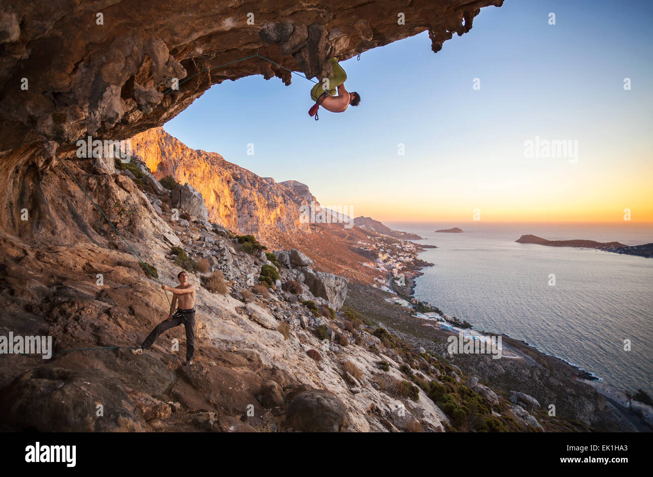 Male rock climber climbing on a roof in a cave, his partner belaying ...