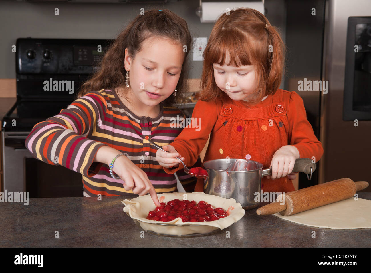 Two sisters adding cherry pie filling to pie crust. Concepts: teamwork ...