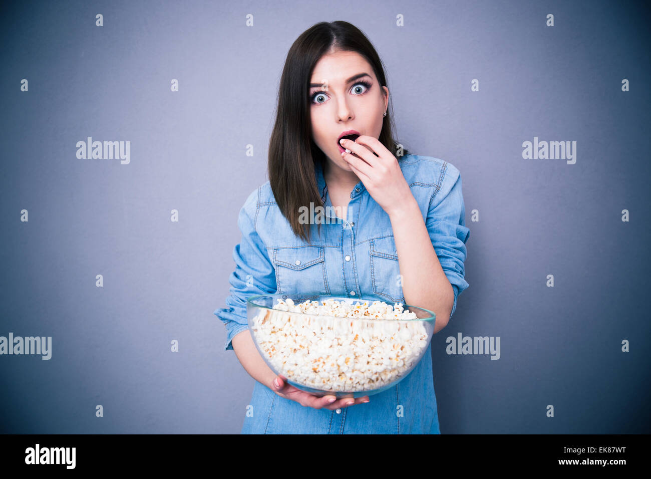 Young surprised woman eating popcorn over gray background. Looking at ...