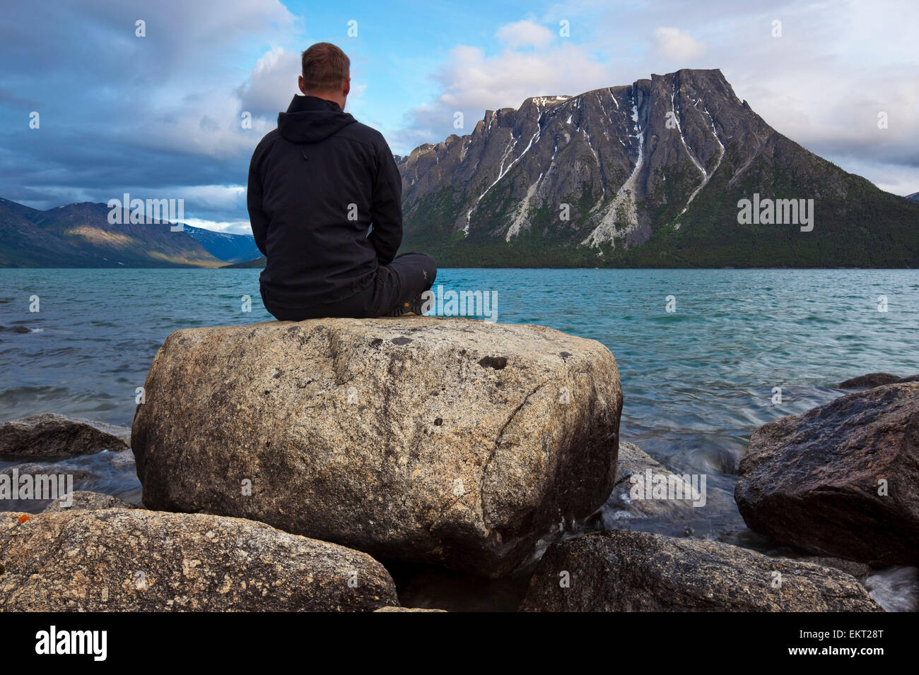 Man sitting on a rock by Kusawa Lake with Ark Mountain in the ...