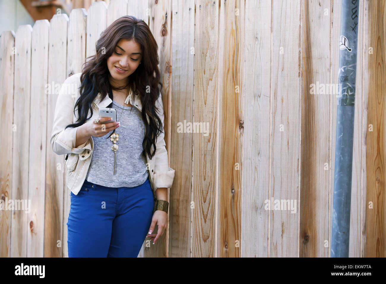 A woman leaning against a wooden fence using her cell phone; San Francisco, California, USA ...