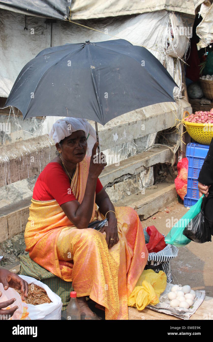 A woman sells produce at Fort Cochin Market, shaded by an umbrella ...