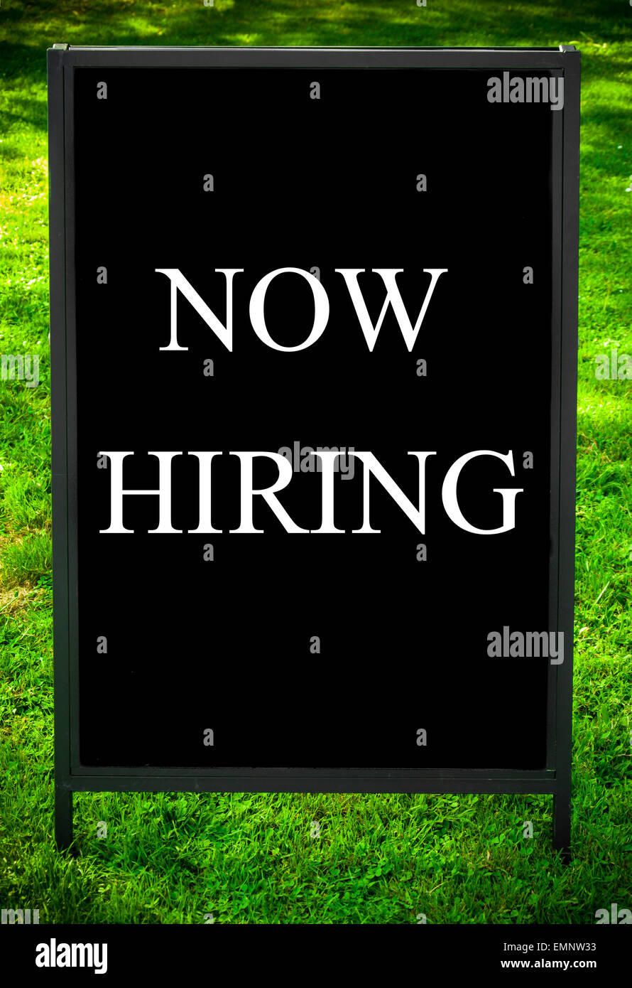 NOW HIRING message on sidewalk blackboard sign against green grass ...