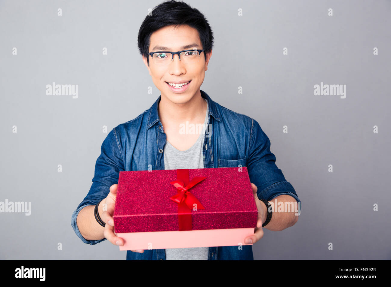 Smiling young asian man giving gift box on the camera over gray ...