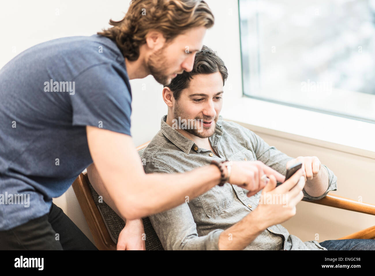 A man seated checking his smart phone and a colleague looking over his ...