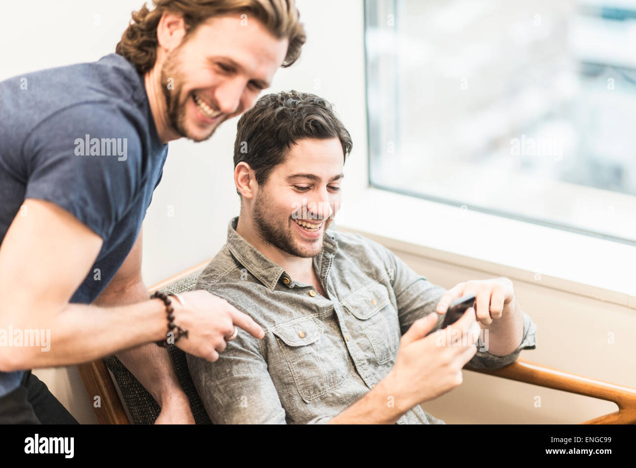 A man seated checking his smart phone and a colleague looking over his ...