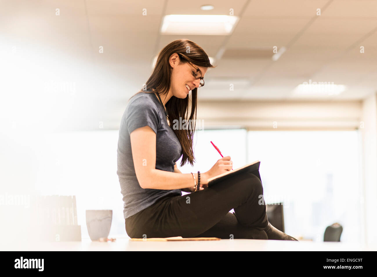 A young woman seated on her desk writing in a notebook with a red pen ...