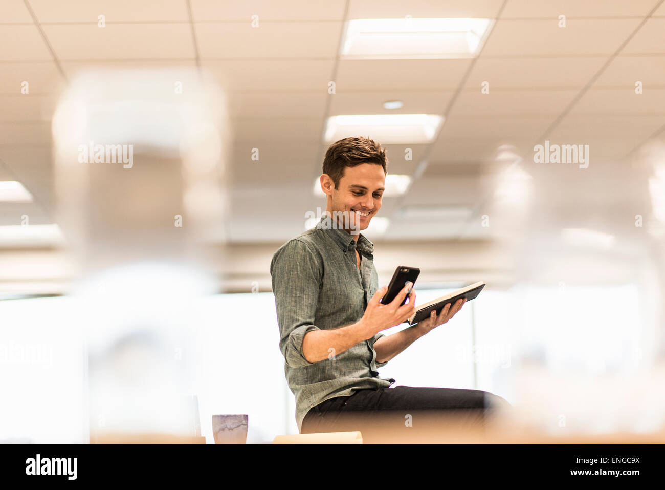 A young man seated looking at his smart phone, holding an open notebook ...