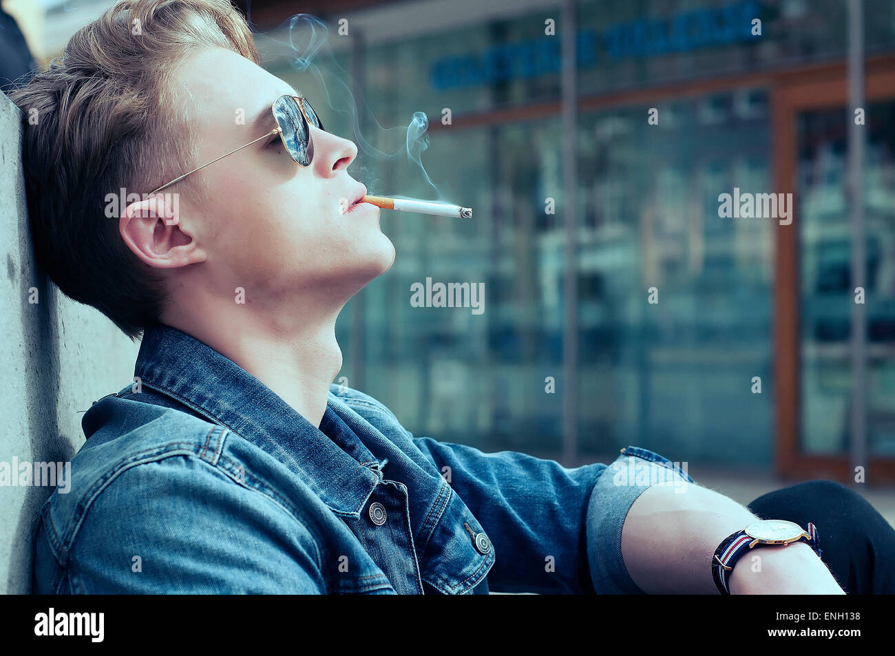 Young Caucasian man in denim jacket and sunglasses smoking a cigarette ...