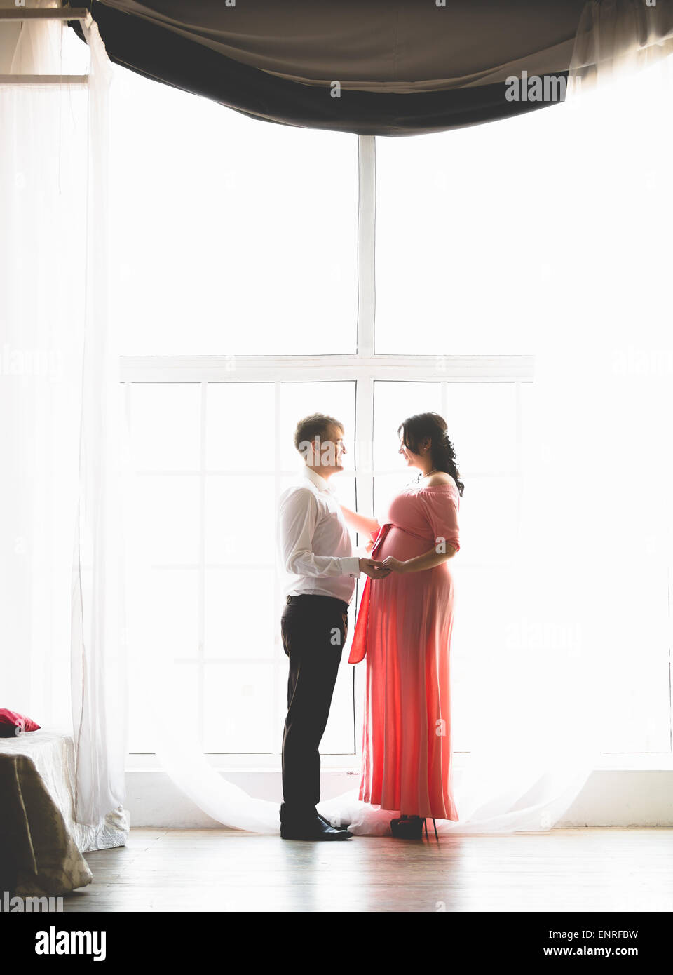 Handsome man and cute pregnant wife standing next to big window at ...