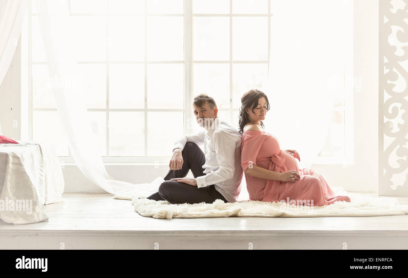 Young pregnant couple sitting back to back on floor against big window ...