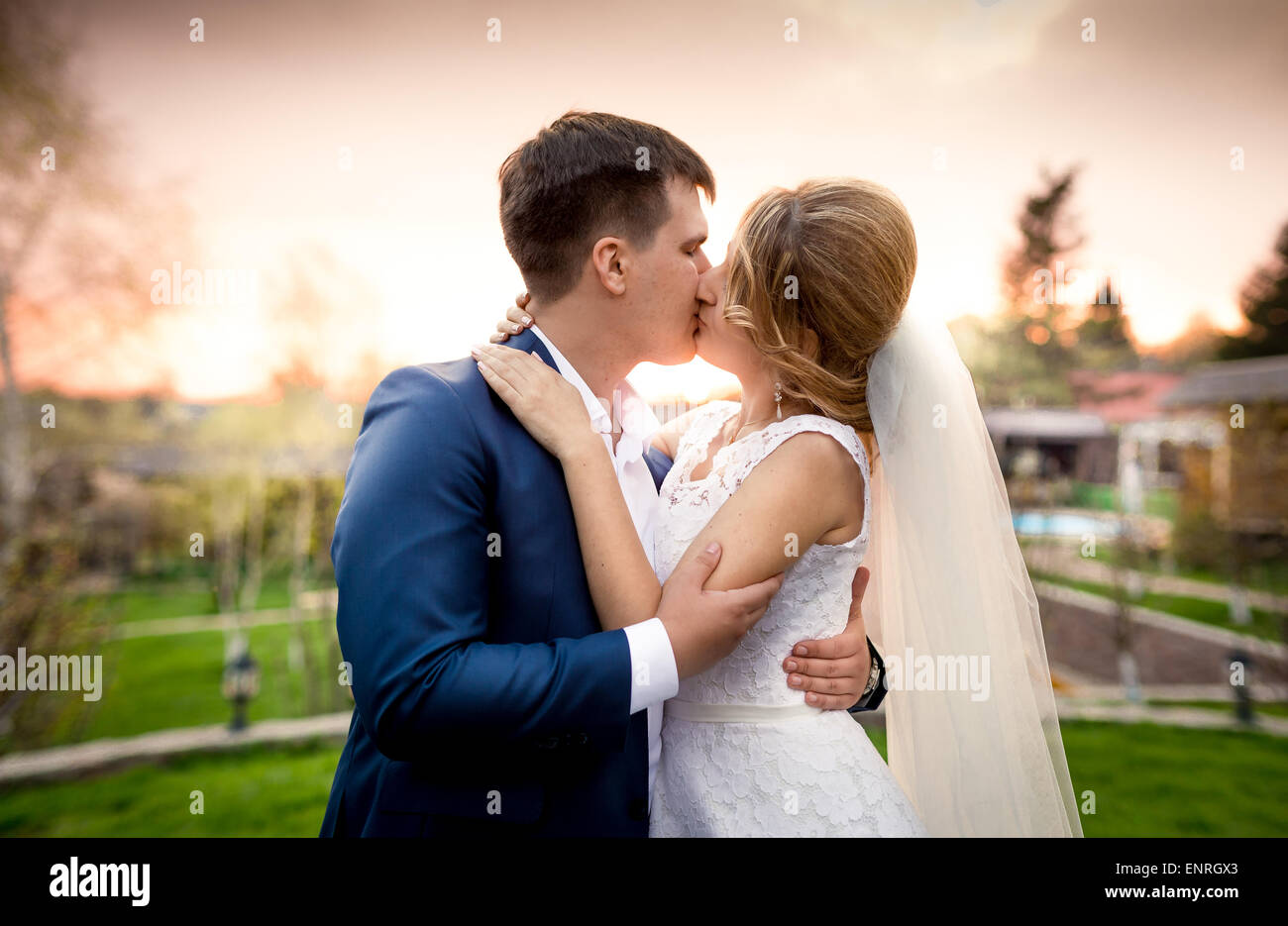 Portrait of elegant newly married couple kissing in park at sunset ...