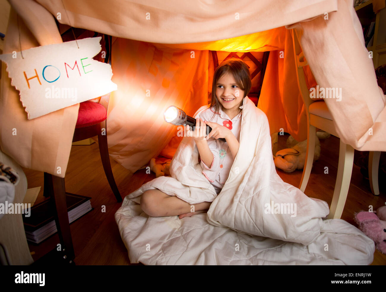 Cute smiling girl playing with flashlight in house made of blankets at ...