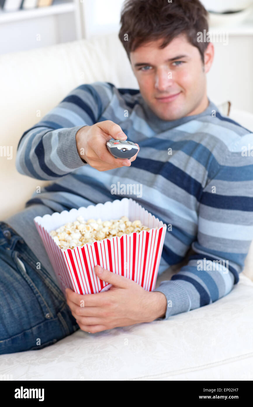 Relaxed young man eating popcorn and holding a remote lying on the sofa ...