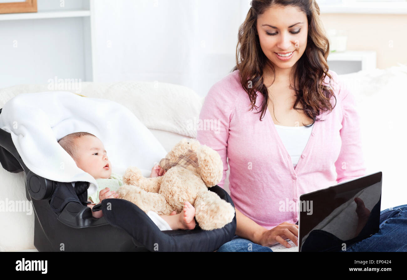 Bright mother working on a laptop with her baby sitting next to her ...