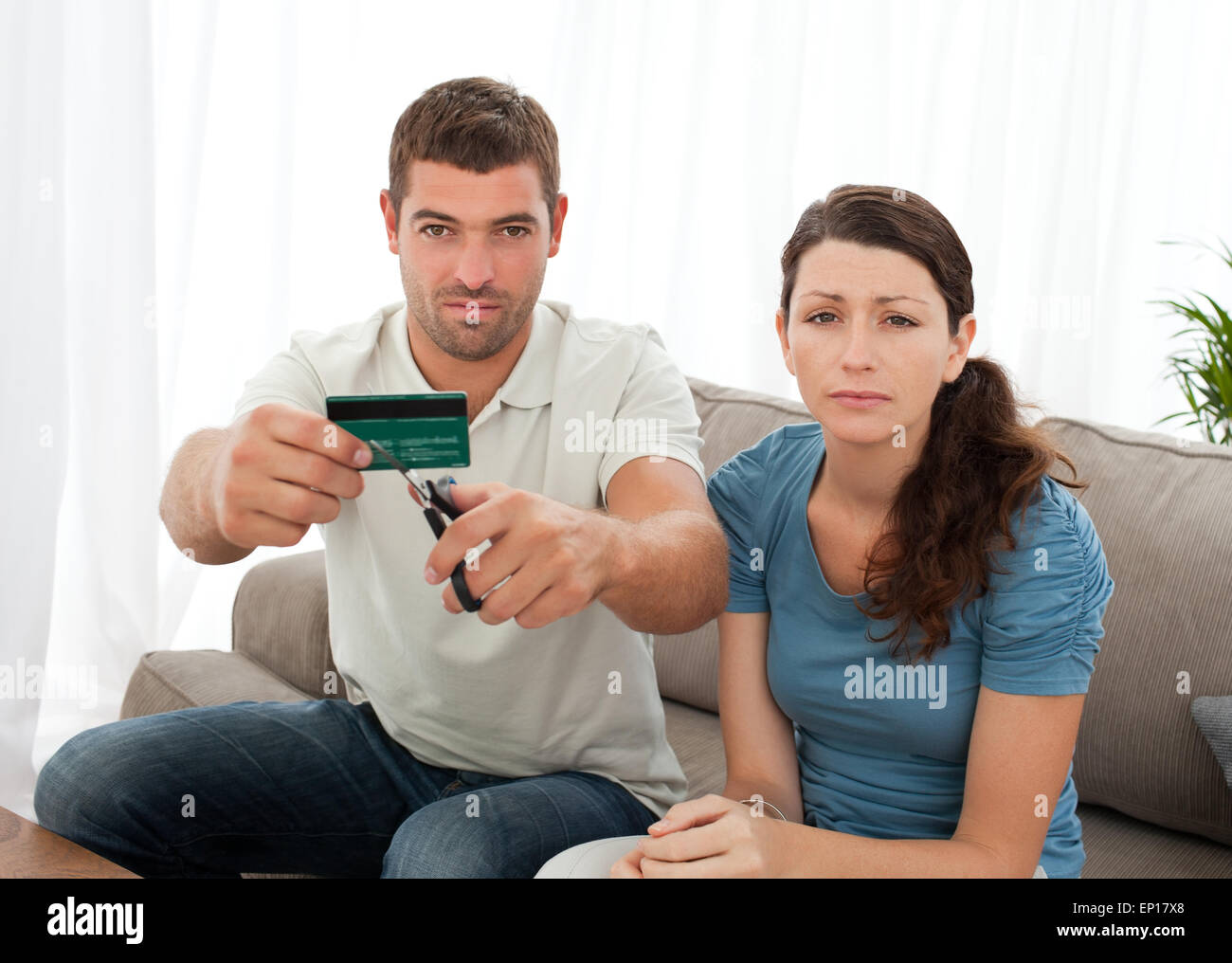 Worried couple cutting their credit card together sitting on the sofa ...