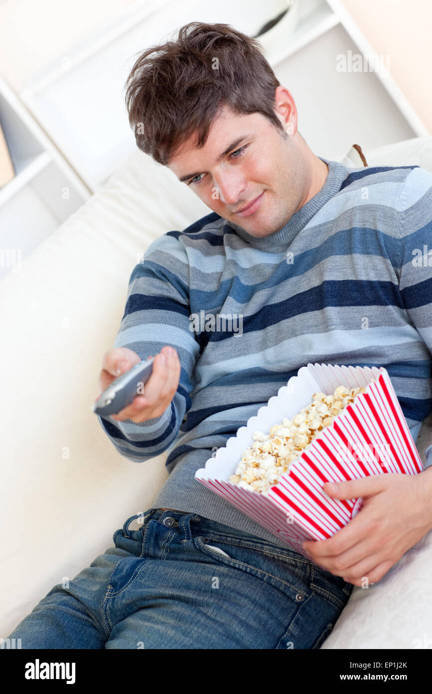 Delighted young man eating popcorn and holding a remote lying on the ...