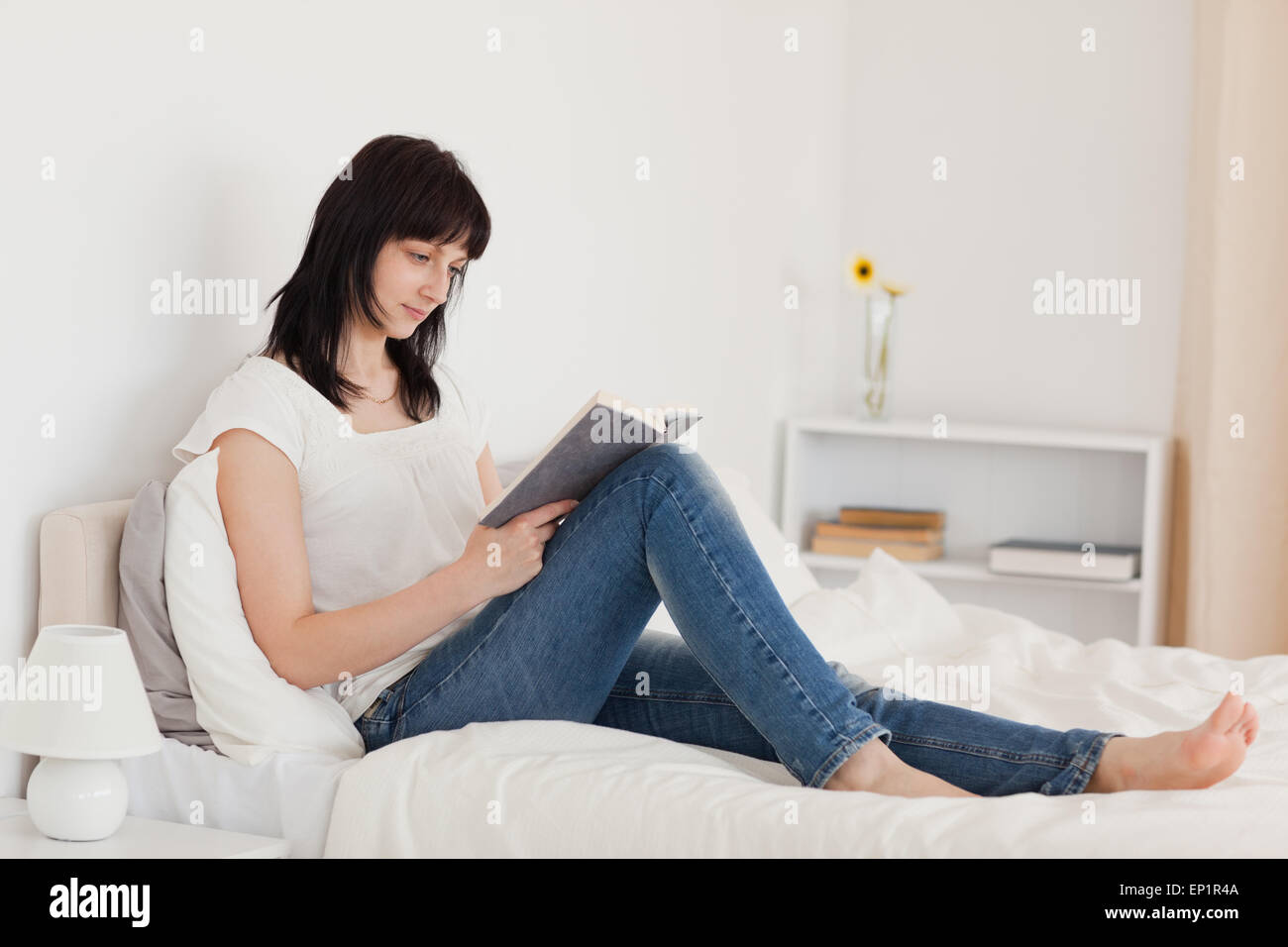 Beautiful brunette woman reading a book while sitting on a bed Stock Photo - Alamy