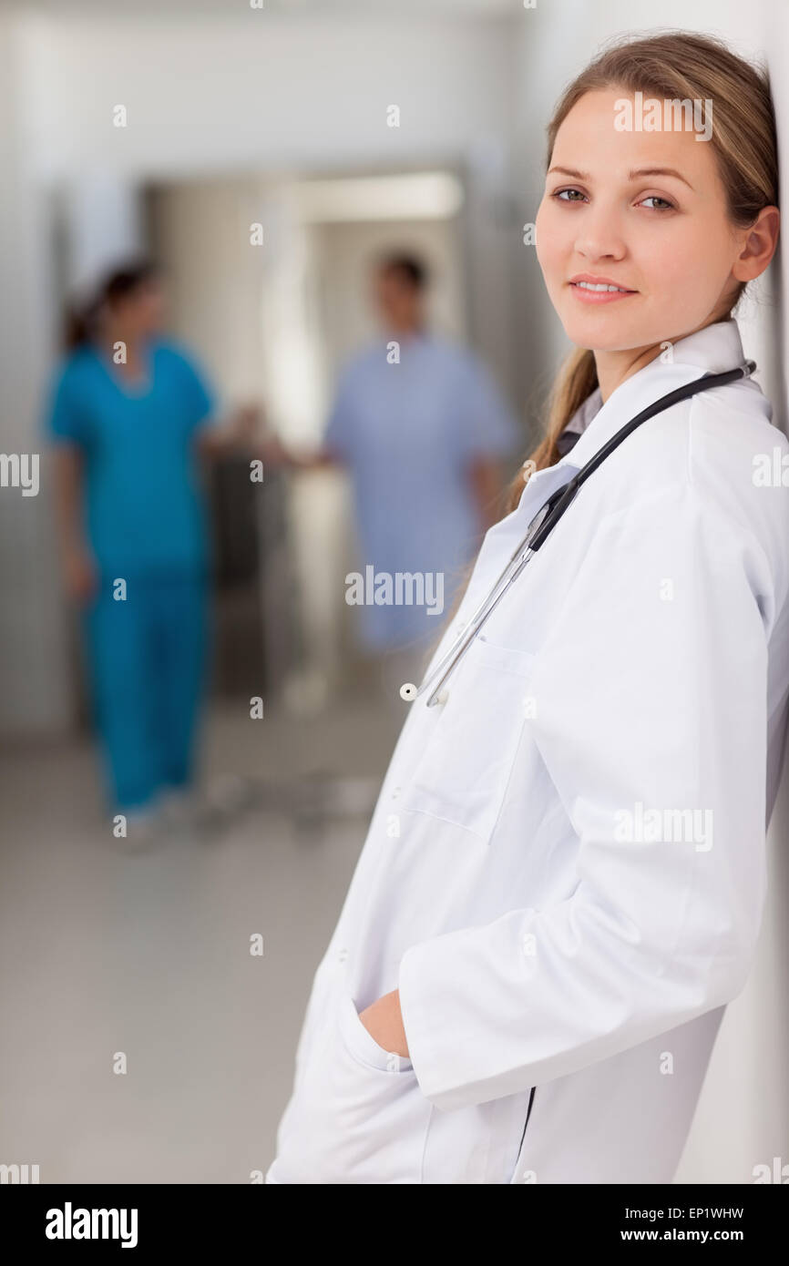 Smiling doctor standing against a wall with her hands in her pockets ...