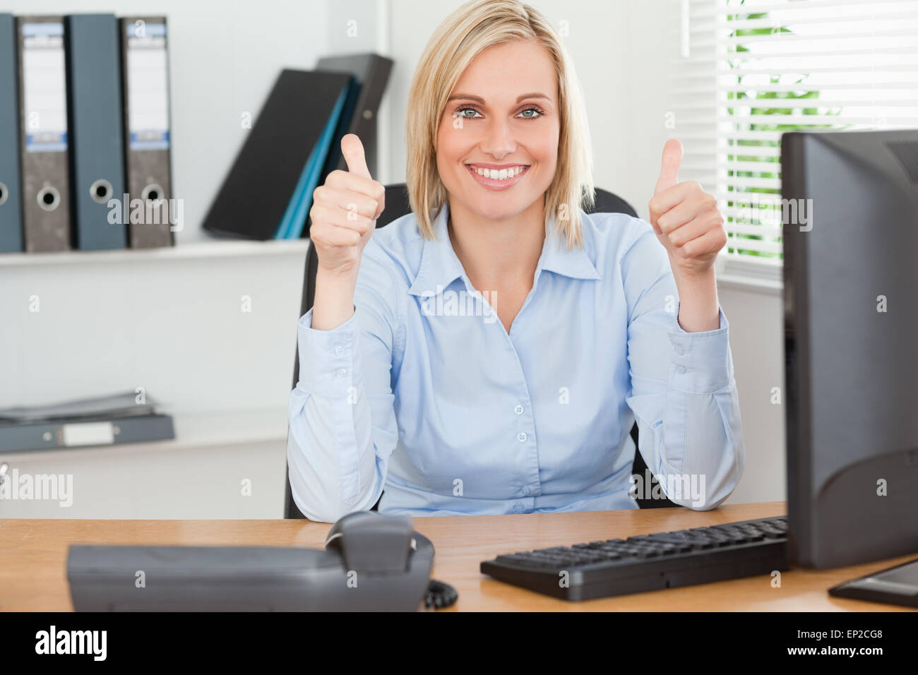 Young woman sitting behind desk with thumbs up Stock Photo - Alamy