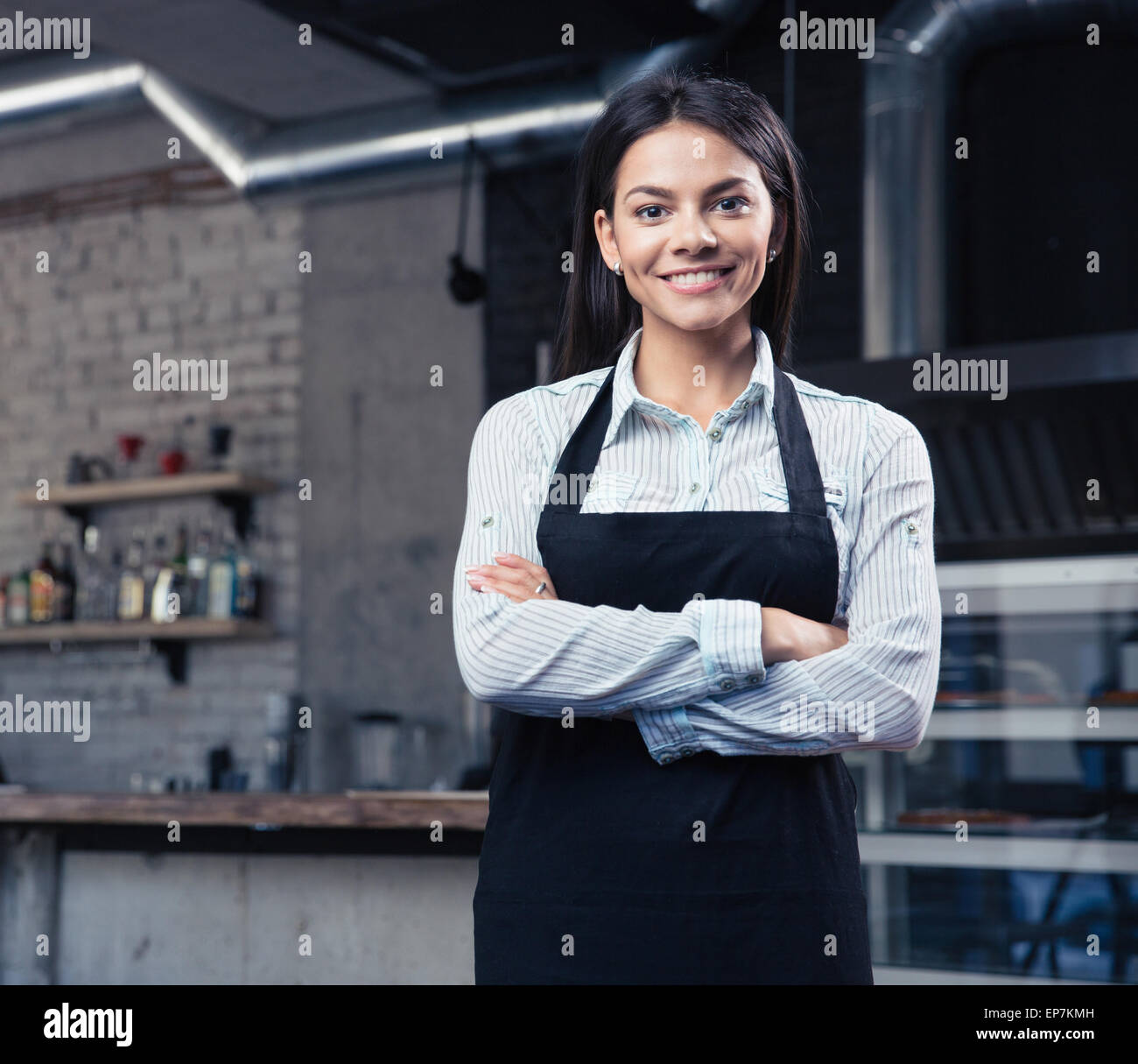 Happy cute female waiter in apron standing with arms folded in cafe ...