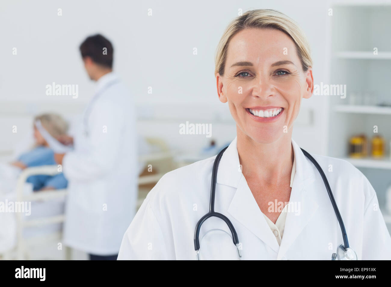 Smiling blond doctor posing with doctor attending patient on background ...