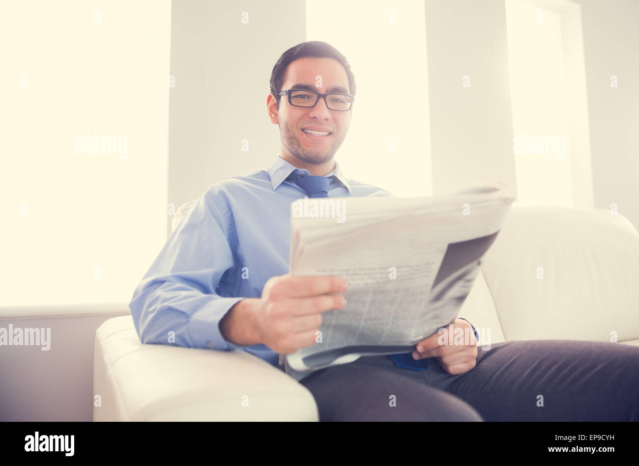 Pleased man looking at camera and holding a newspaper sitting on a sofa ...