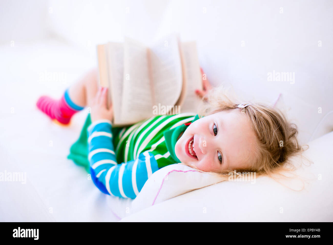 Happy laughing little girl reading a book relaxing on a white couch at ...