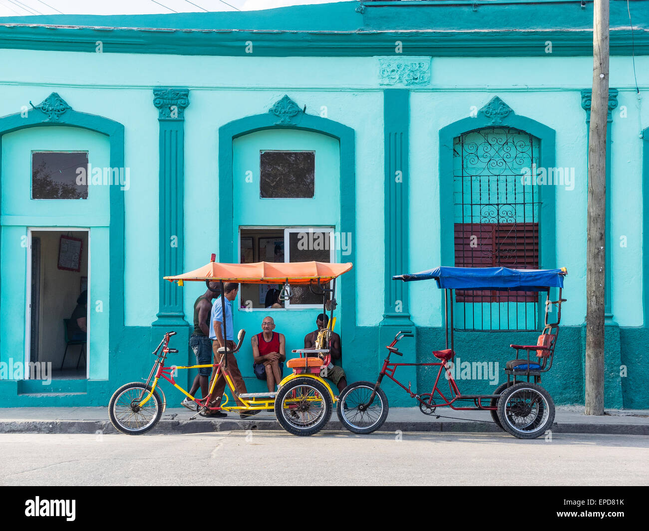 Cuban pedicab drivers wait beside a colorful building by their pedicabs ...