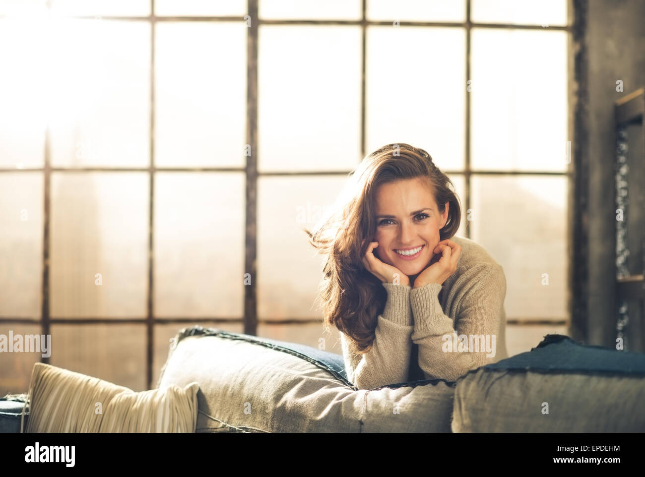 A brunette woman with long brown hair leans over a sofa, smiling ...