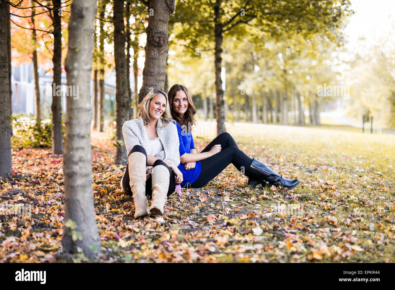 Two female friends leaning on tree trunk Stock Photo - Alamy