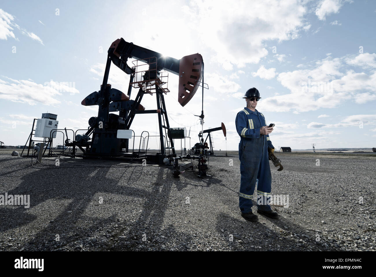 A man in overalls and hard hat at a pump jack in open ground at an oil ...