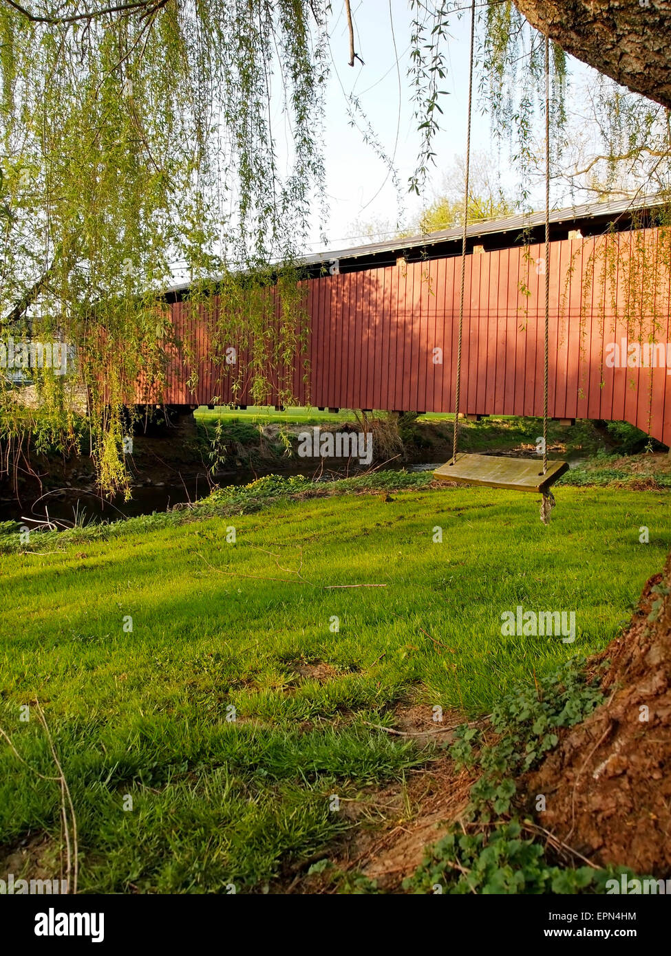A tree swing hangs from a willow tree near a red covered bridge over a ...