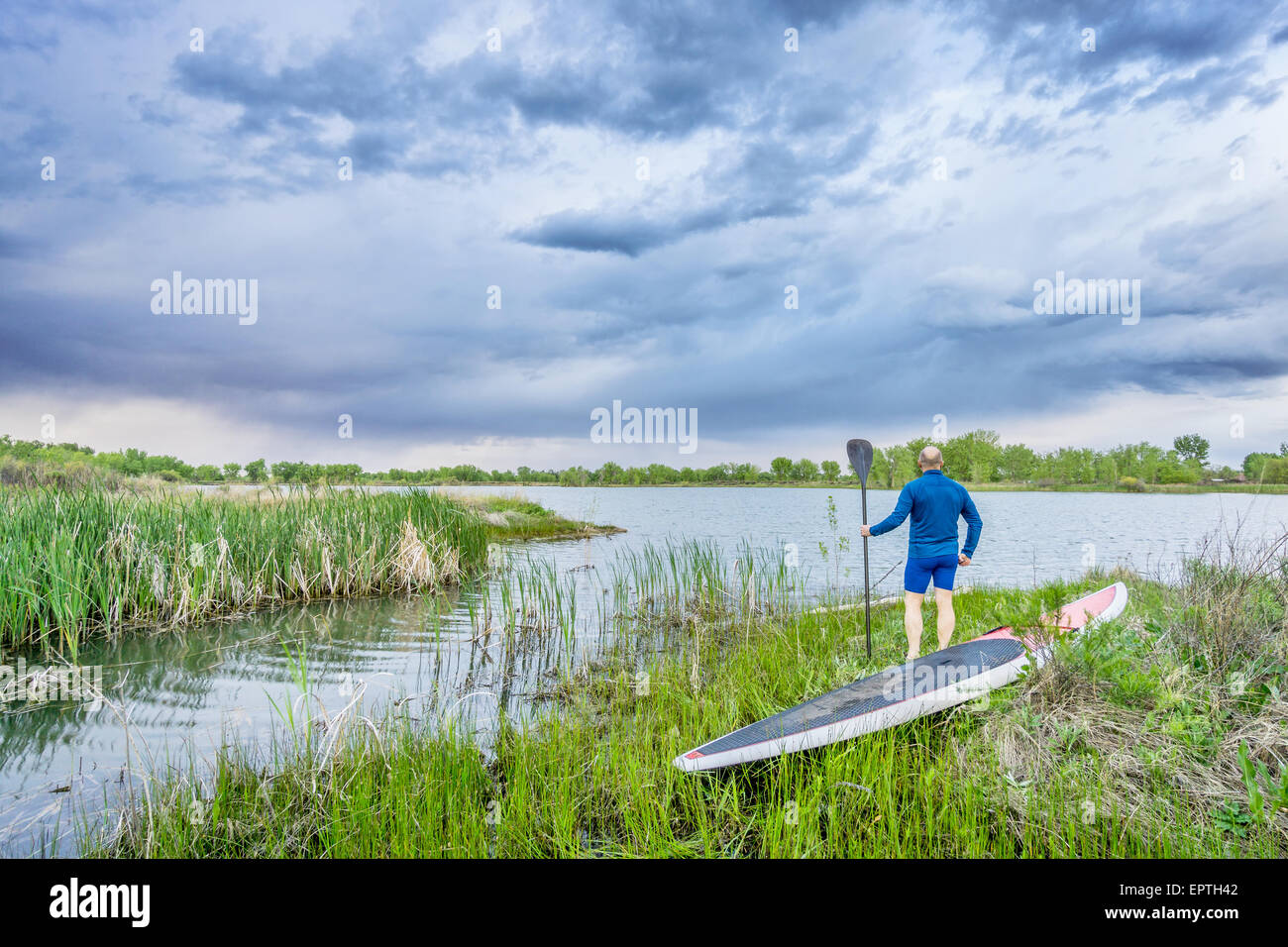 senior male with stand up paddleboard on lake shore watching stormy ...