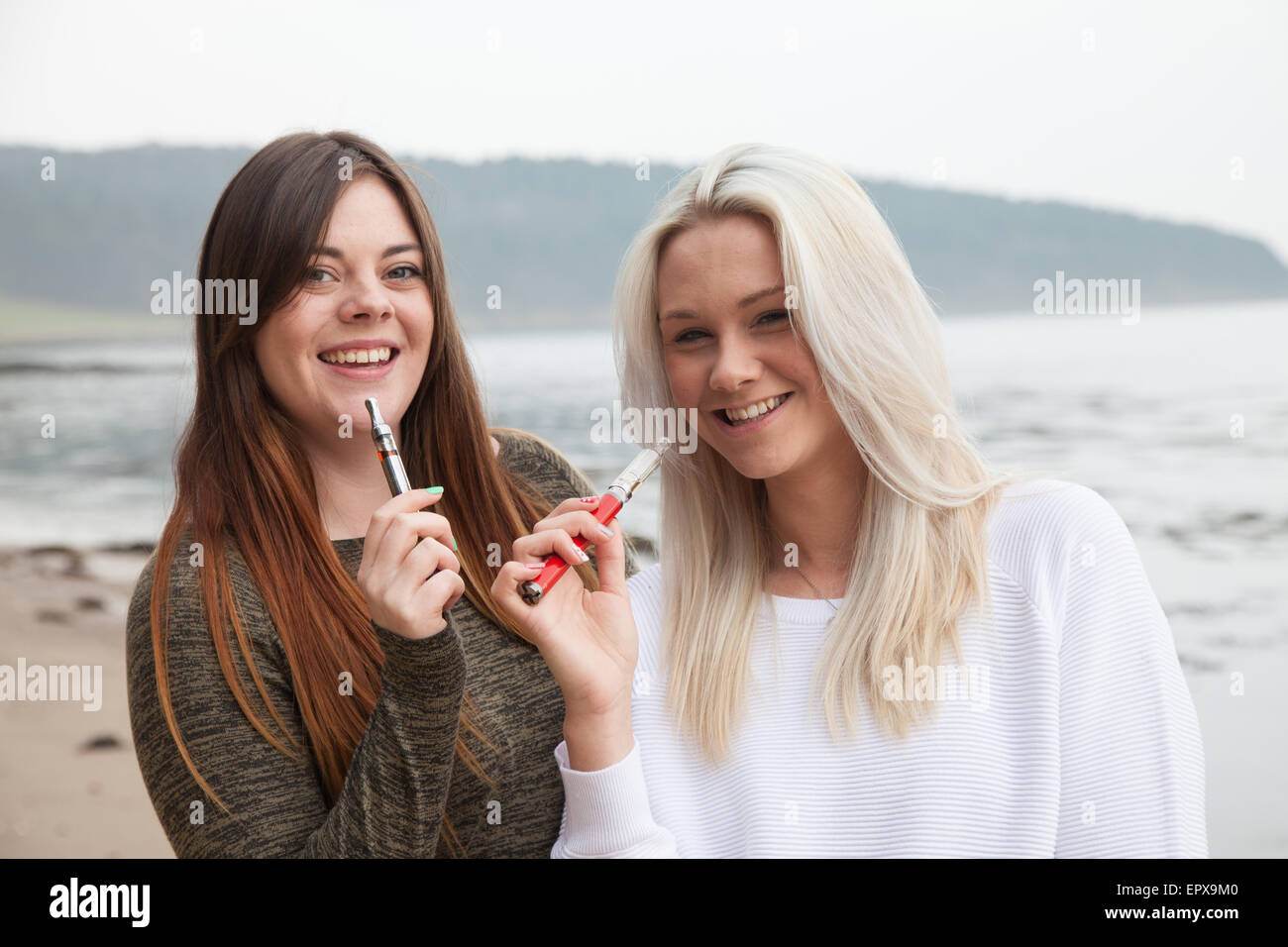 Two women in their 20's holding personal vaporizer's outside on a beach ...