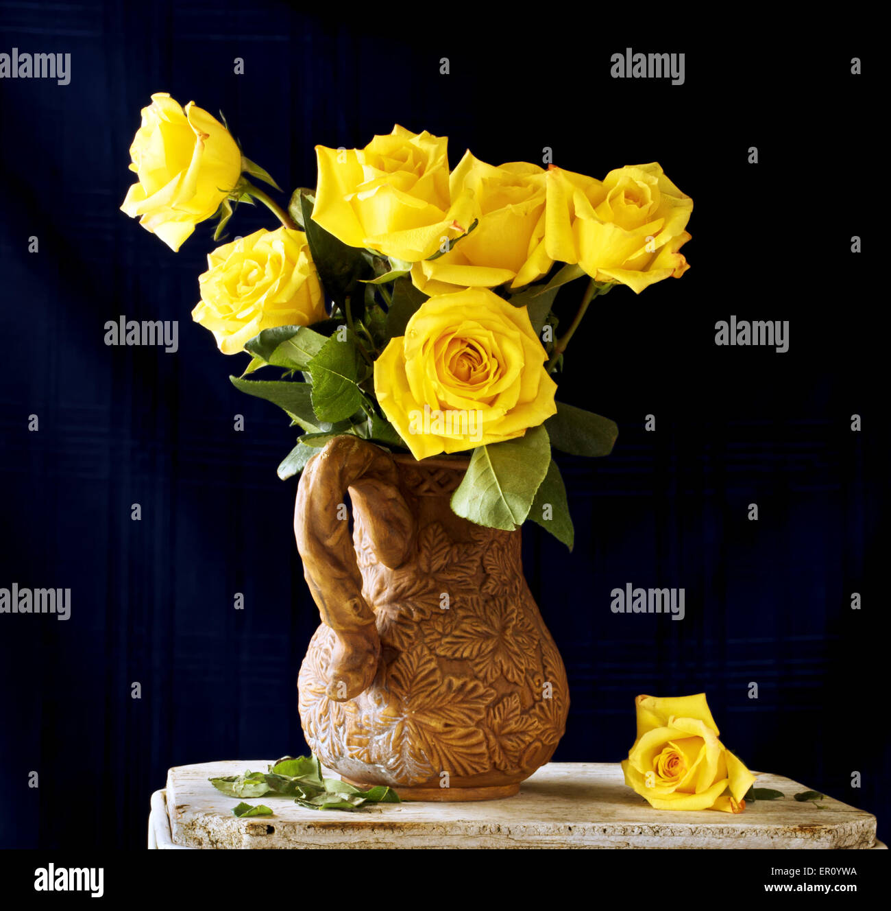 Yellow roses in a brown water pitcher on a distressed white table. Dark ...